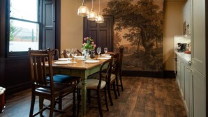 Uma área de refeições com uma mesa de madeira posta para seis pessoas, rodeada por cadeiras. Um grande mural com tema de árvores decora a parede atrás da mesa.
