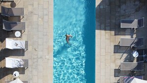 Vista aérea de uma piscina retangular com uma pessoa nadando, rodeada por espreguiçadeiras e guarda-sóis.