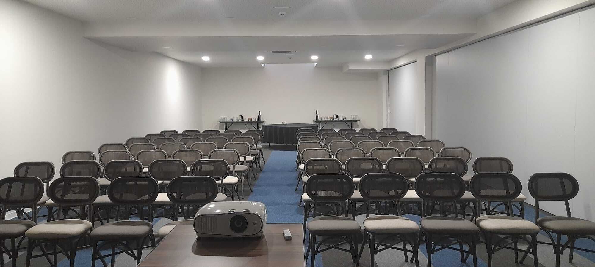 Uma sala de conferências com fileiras de cadeiras, um projetor sobre uma mesa e uma mesa na frente.