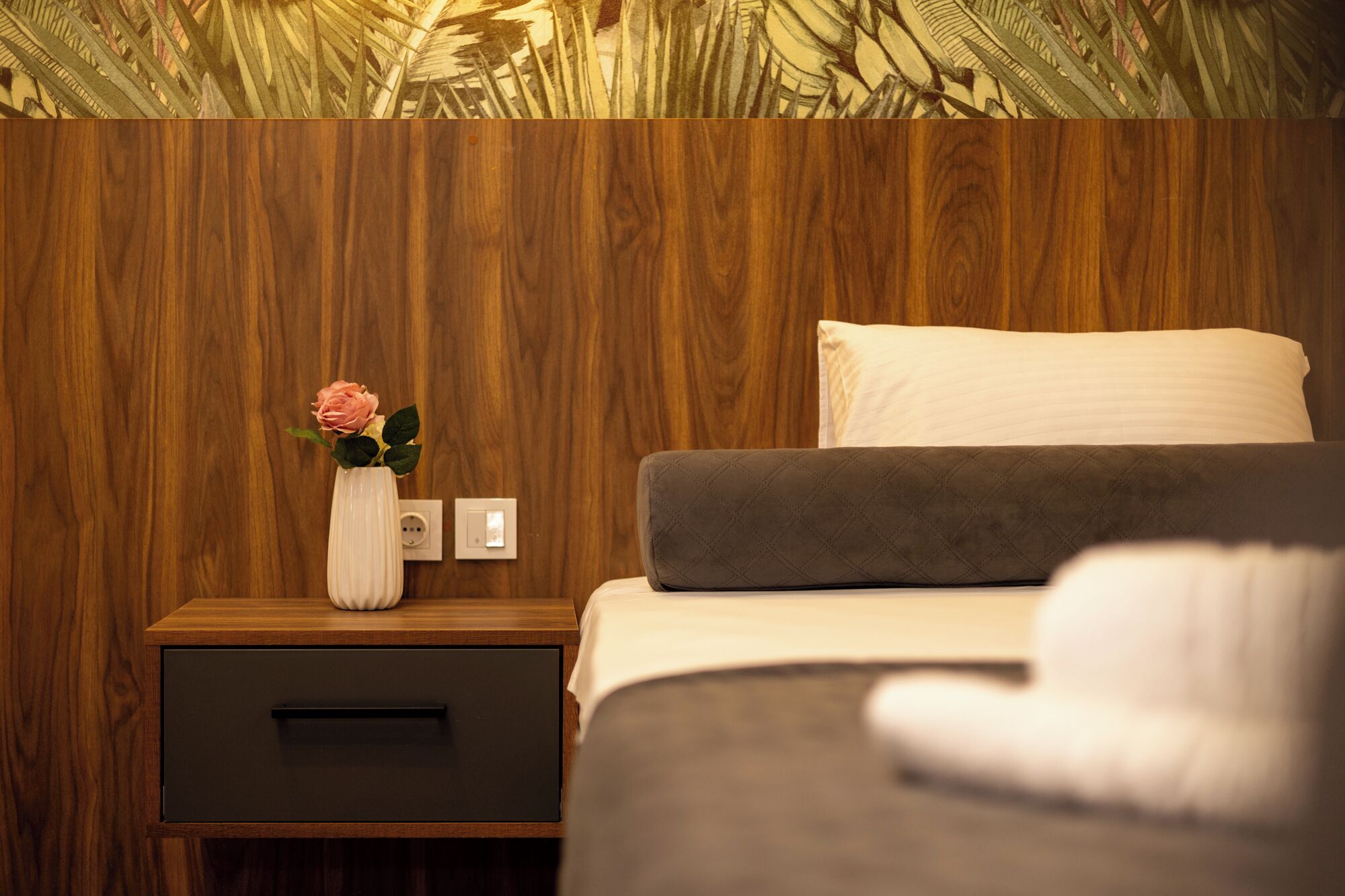Quarto com cabeceira de madeira, mesa de cabeceira com um vaso de flores, cama com travesseiro e gaveta lateral.