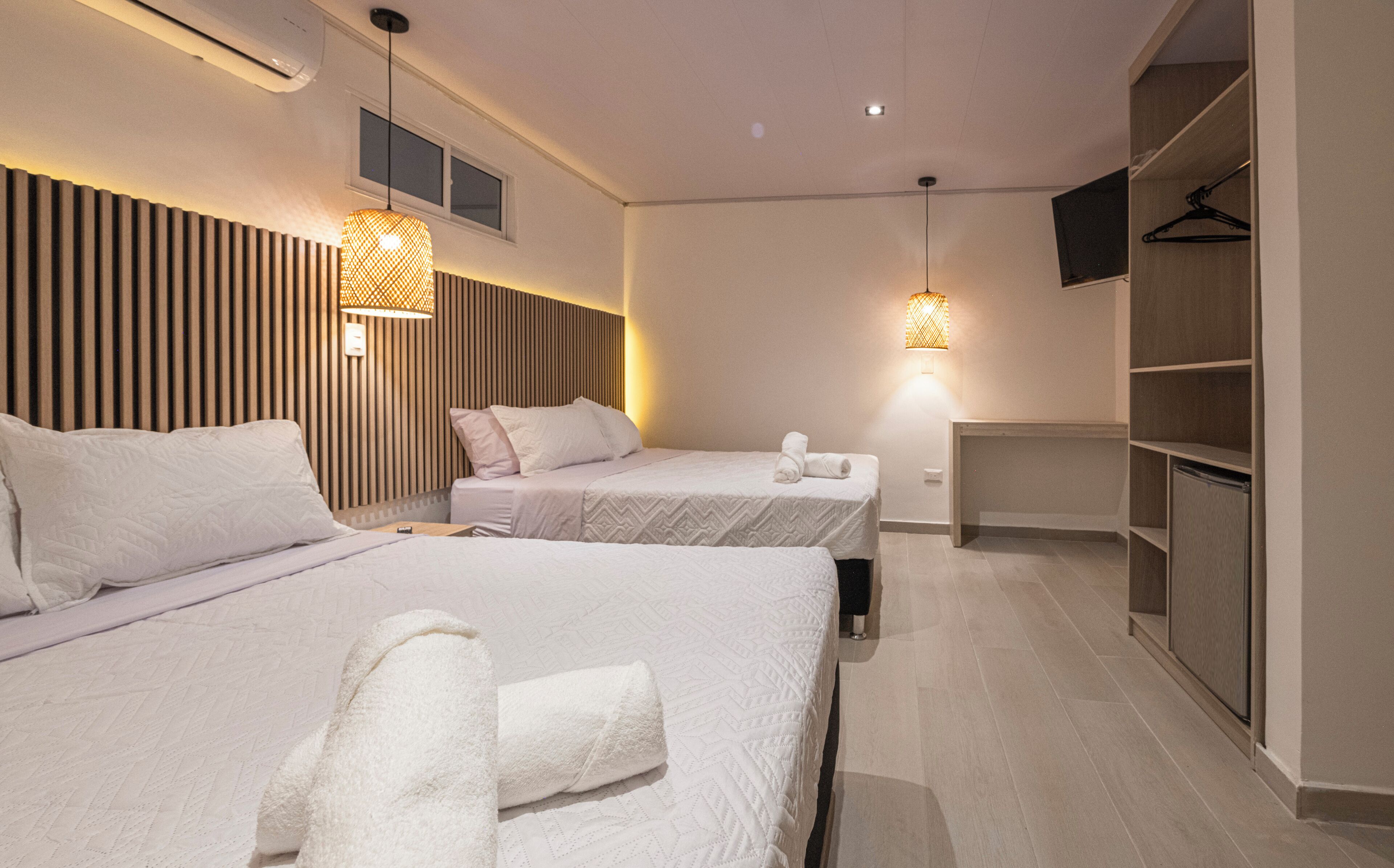 Quarto de hotel com duas camas, televisão, guarda-roupa e mesa de cabeceira.