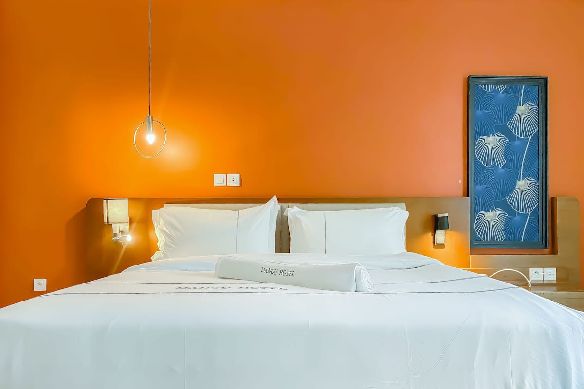 Quarto de hotel com cama grande, roupa de cama branca, luminária pendente, obra de arte na parede e mesa de cabeceira com abajur.