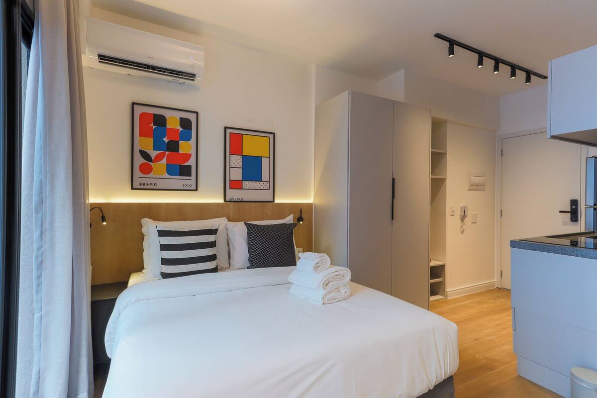 Quarto de hotel moderno com uma cama, duas obras de arte abstrata emolduradas, ar condicionado integrado, abajur e uma pequena cozinha.
