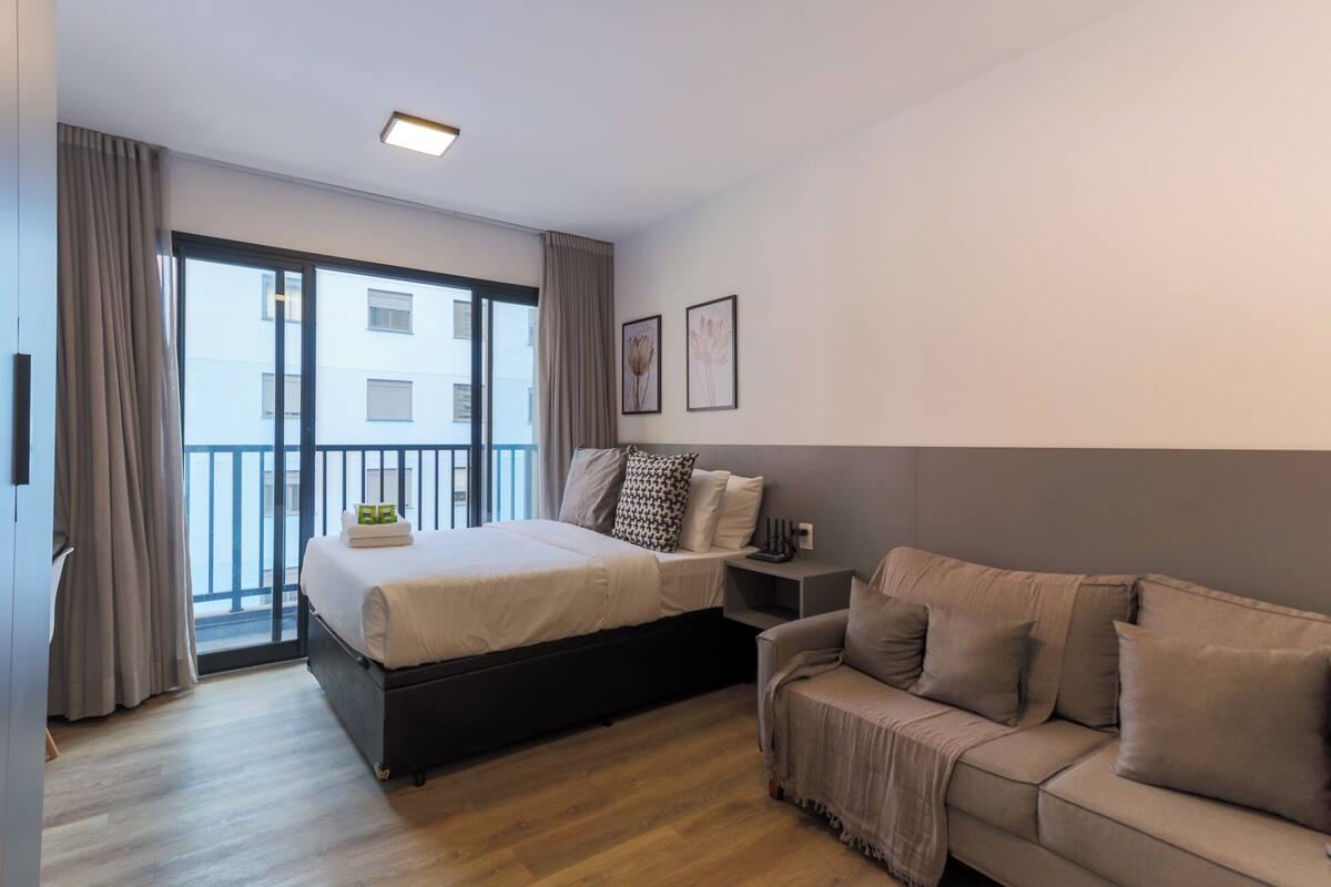 Quarto de hotel moderno com cama, sofá, pequena mesa e varanda com cortinas.
