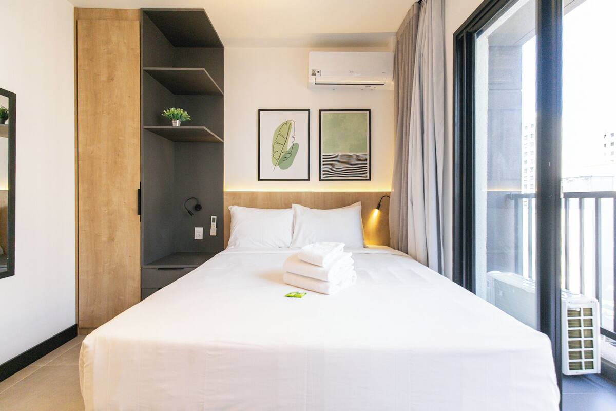 Quarto de hotel com uma cama grande, um guarda-roupa de madeira, uma pequena planta, duas obras de arte emolduradas e uma janela com cortinas.