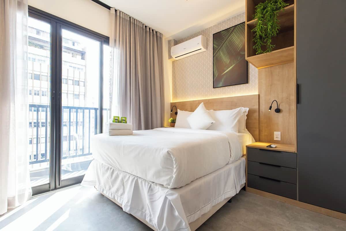 Quarto de hotel moderno com uma cama grande, uma mesa de cabeceira, ar condicionado fixado na parede, uma planta e vista da cidade pela janela.