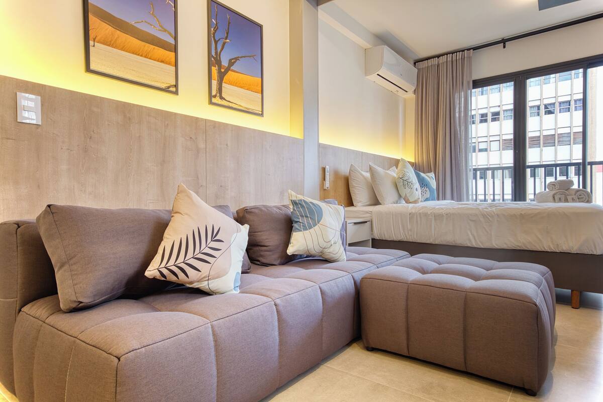 Quarto moderno com sofá-cama, uma cama separada e uma grande janela com vista para prédios.