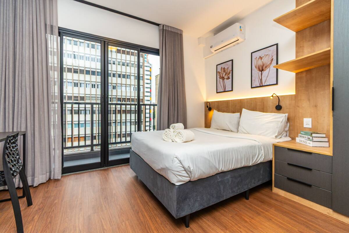 Quarto de hotel moderno com uma cama grande, uma escrivaninha, uma cadeira e vista para o horizonte urbano através de uma janela ampla.