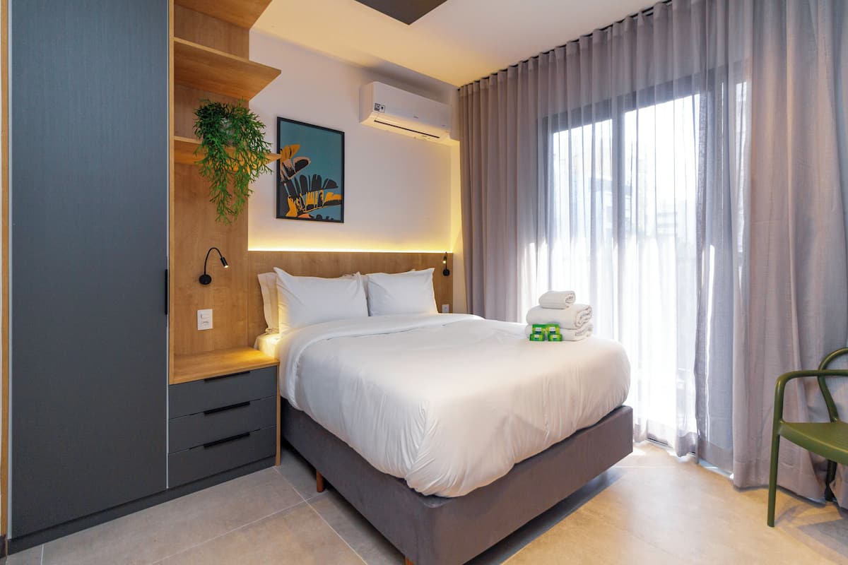 Quarto moderno com cama grande, mesa de cabeceira, ar condicionado fixado na parede, planta em vaso e janela com cortinas.