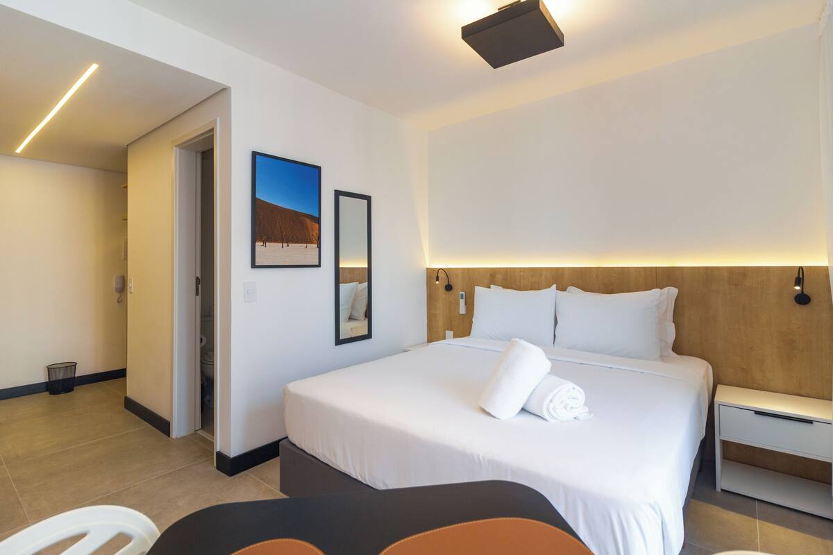Quarto de hotel com uma cama grande, mesa de cabeceira, cadeira e um quadro na parede.