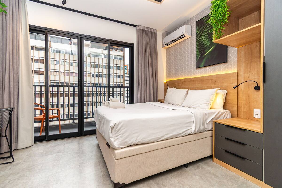 Quarto moderno com uma cama grande, mesa de cabeceira de madeira, escrivaninha e vista para uma varanda através de uma porta deslizante.