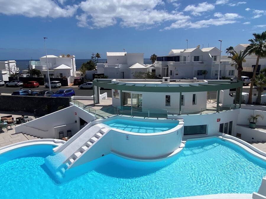 Um hotel moderno com piscina central, edifícios brancos e vista para o mar.