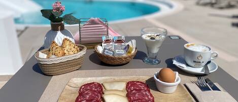 Mesa com diversos alimentos, incluindo pão, queijo, salame e uma xícara de café, com vista para a piscina.