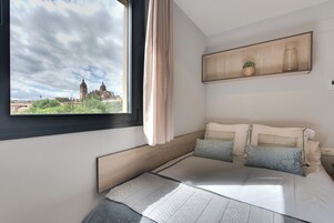 Quarto com uma grande janela, uma cama com roupa de cama estampada, uma cabeceira de madeira, uma estante com uma planta e um pote, e vista para um edifício histórico.