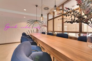 Uma sala de conferência com uma mesa de madeira comprida, cadeiras azuis, luminárias pendentes e um letreiro de neon na parede.