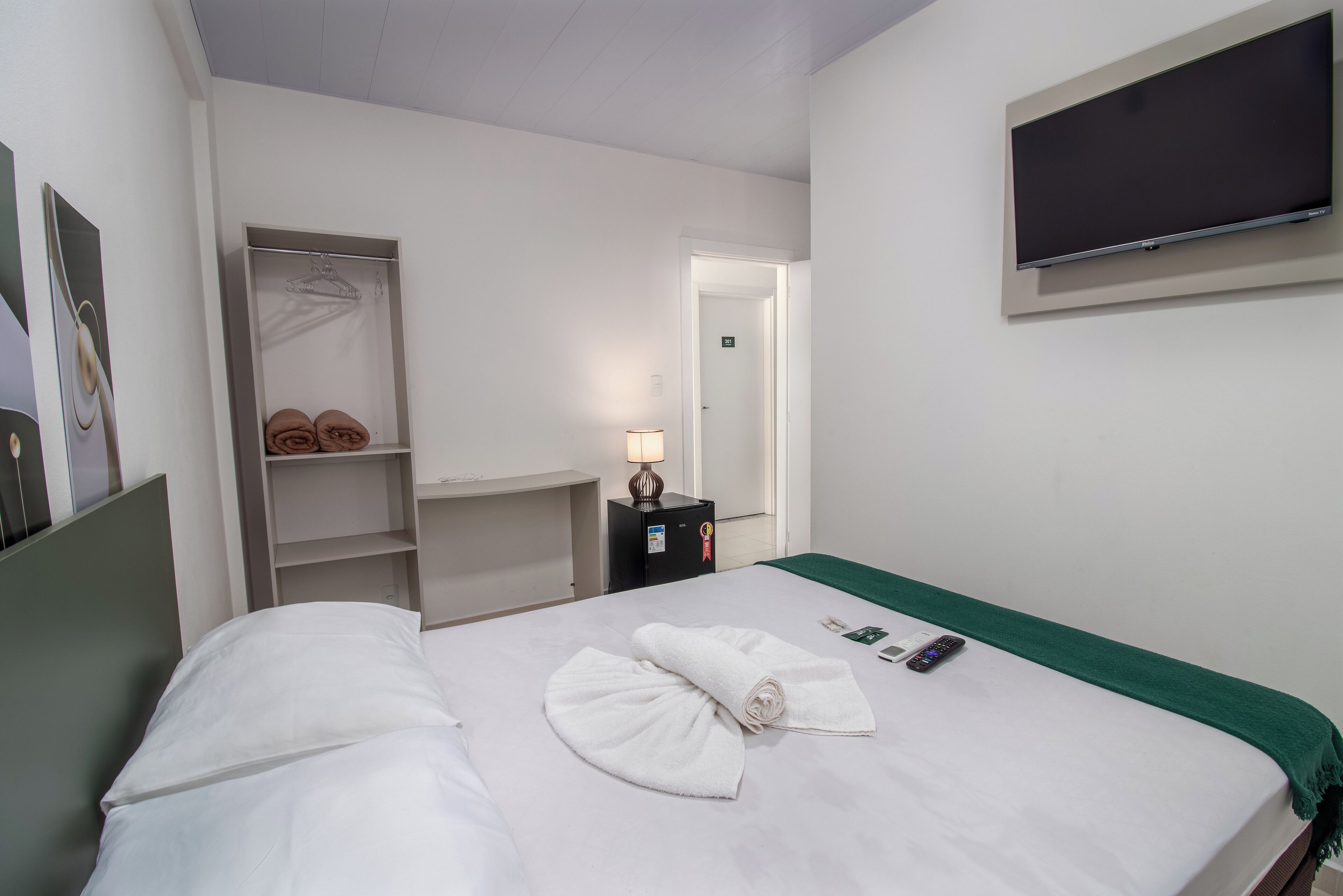 Quarto de hotel com cama, televisão na parede, guarda-roupa e mesa de cabeceira com abajur.