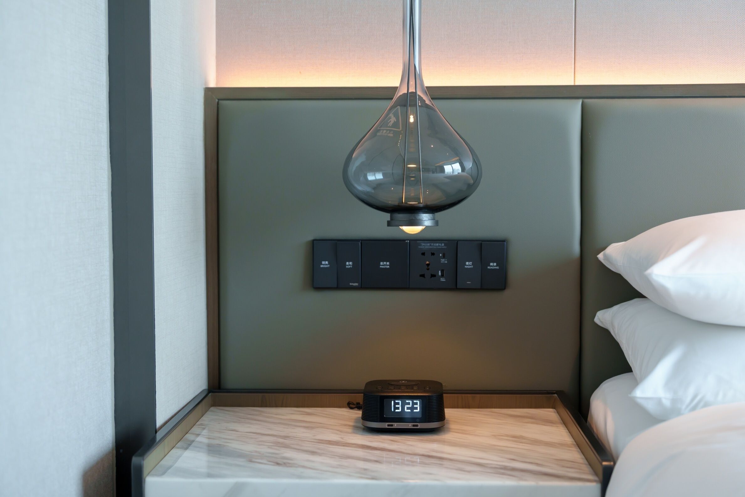 Quarto de hotel moderno com uma mesinha de cabeceira, um relógio digital mostrando a hora, uma tomada na parede e um candeeiro suspenso.