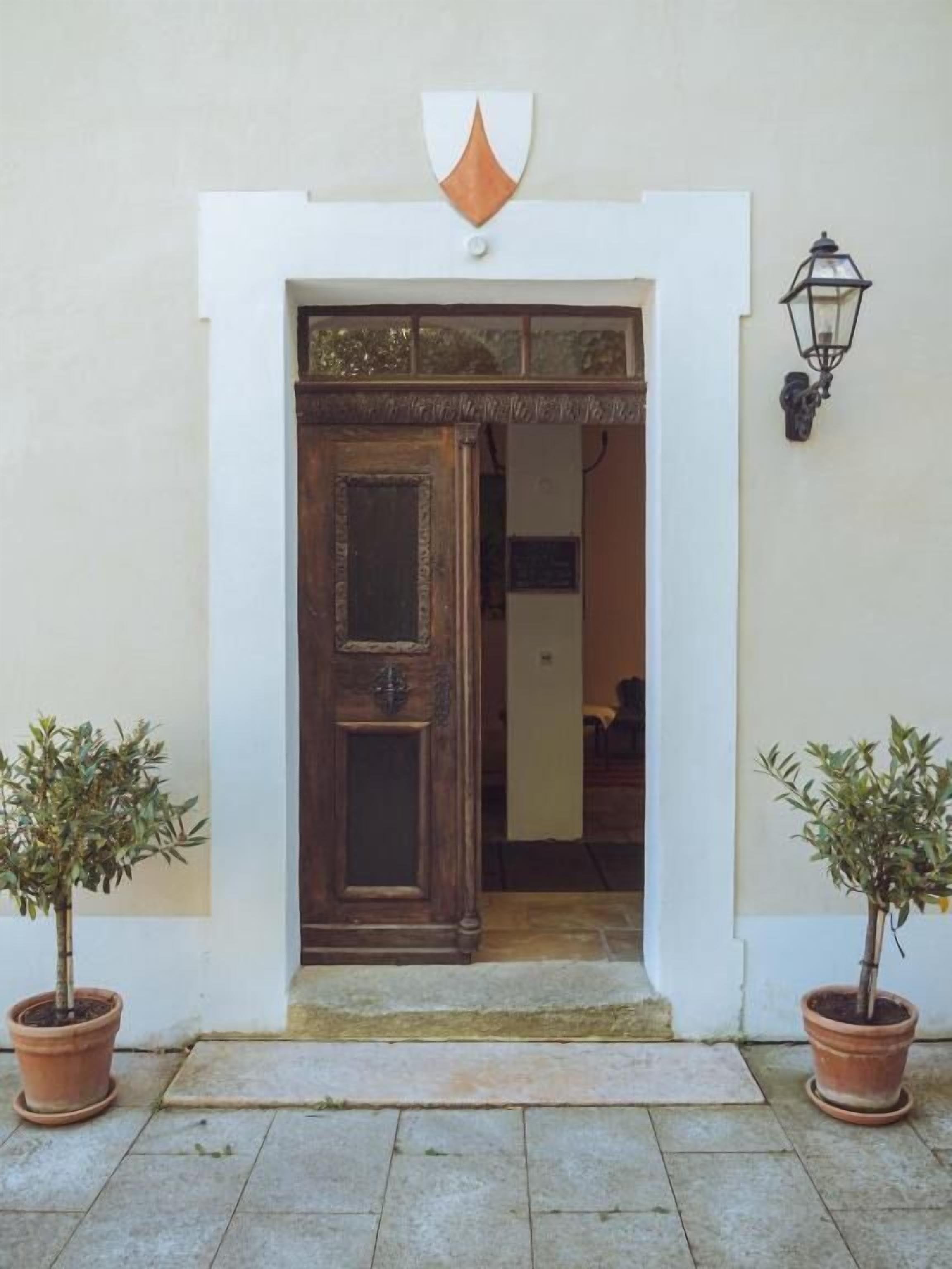 Uma porta de madeira com detalhe decorativo na parte superior, emoldurada por dois vasos de plantas, dando acesso a um espaço interno.