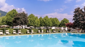 Piscina com espreguiçadeiras e guarda-sóis, cercada por árvores e edifícios.