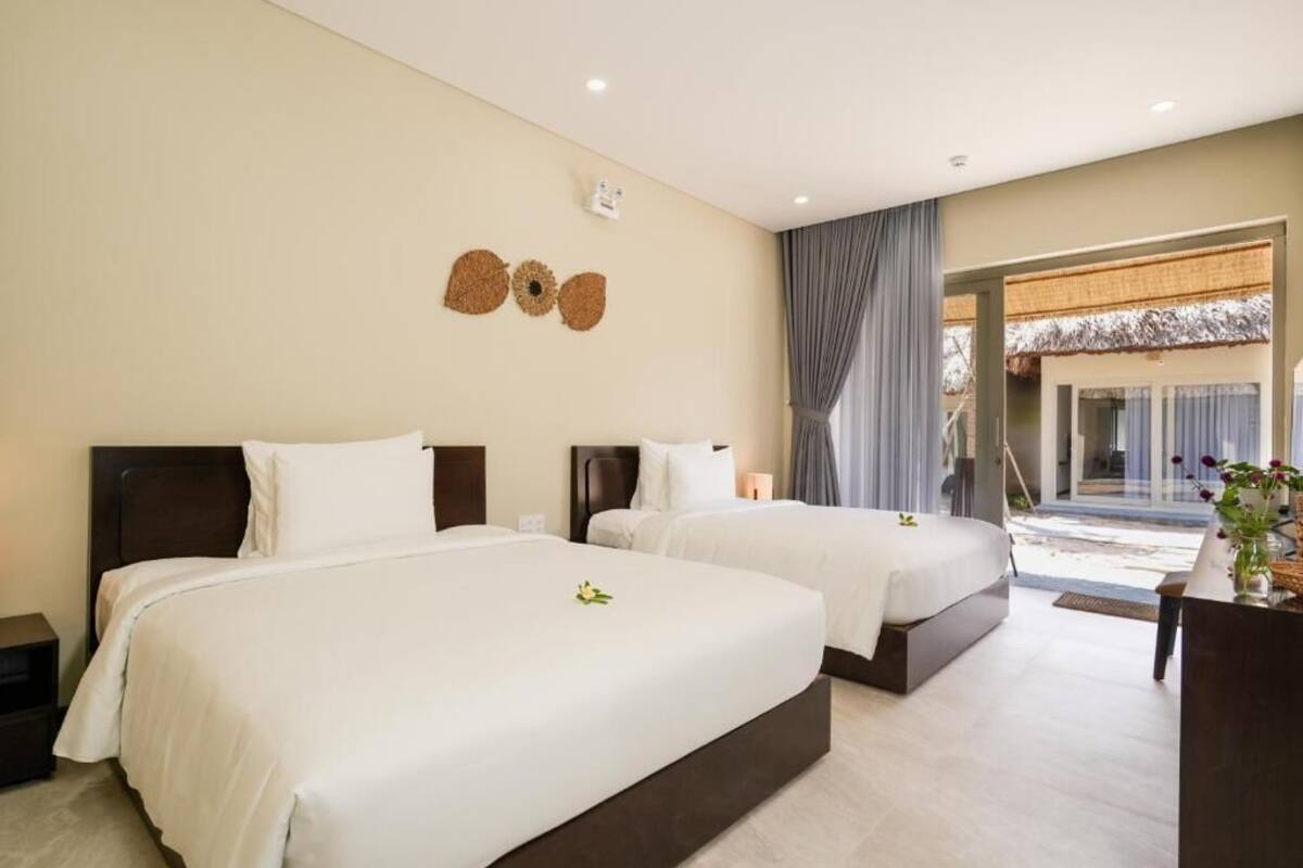 Quarto de hotel com duas camas, uma janela grande com cortinas e vista para uma construção com telhado de palha.