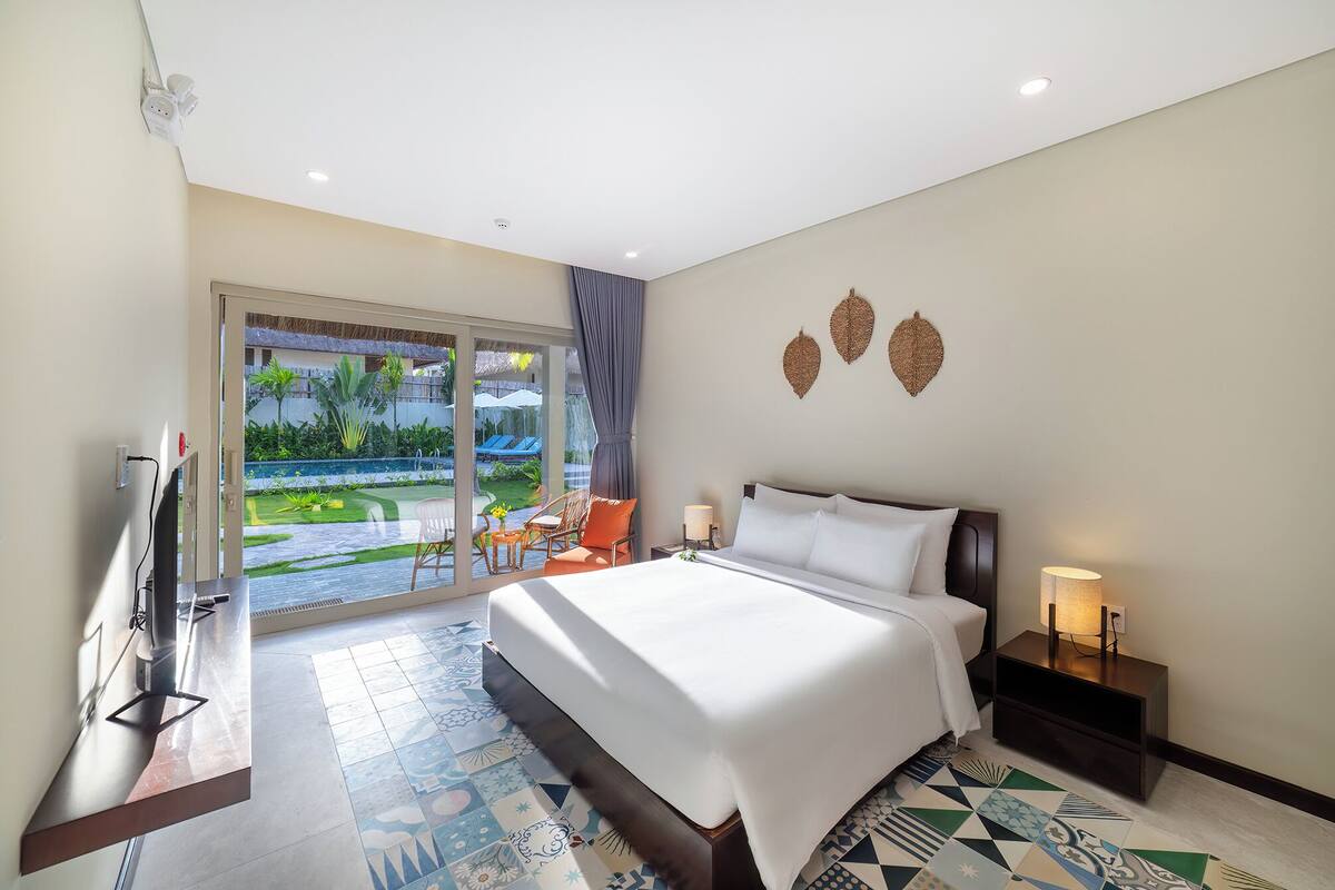 Quarto de hotel com cama, televisão, mesa de cabeceira e vista para a piscina e jardim.