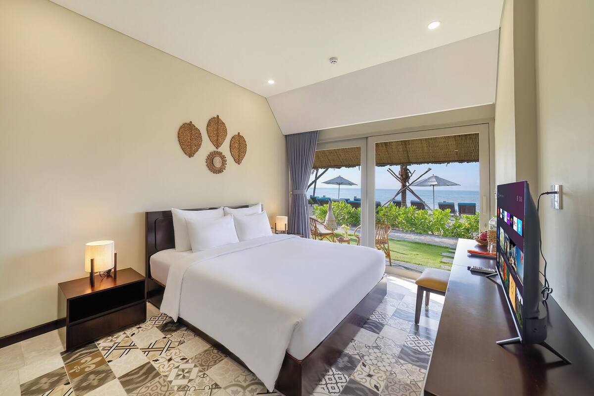 Quarto de hotel com uma cama grande, uma escrivaninha com uma TV e vista para o mar através de amplas janelas.