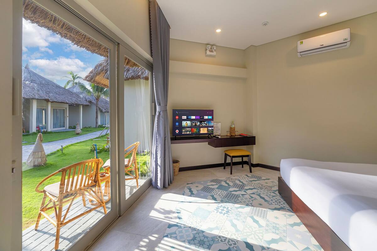 Quarto de hotel com uma grande porta de correr de vidro que dá para uma área externa privativa, com uma estrutura de telhado de palha e um gramado.