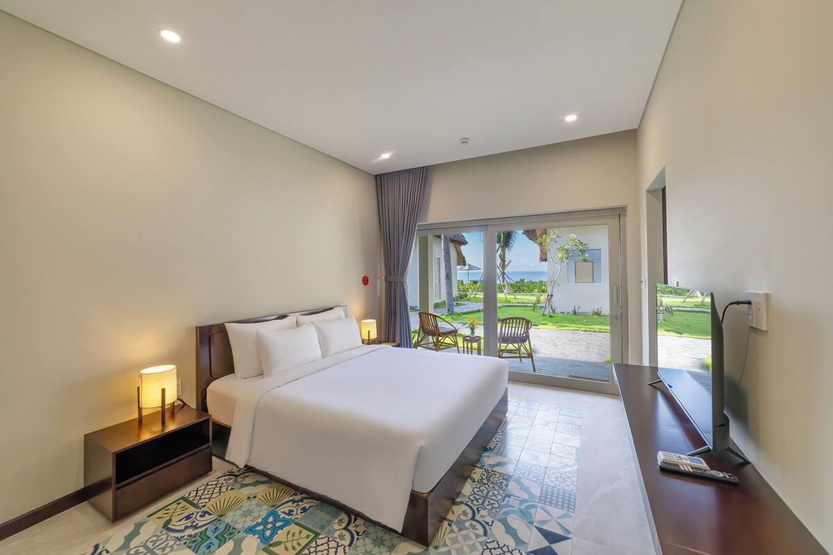 Quarto de hotel moderno com uma cama grande, televisão, secretária e vista para o jardim e piscina externos.