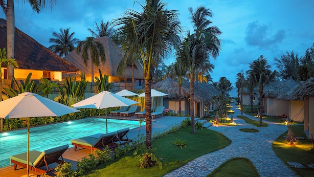 Área de piscina de resort com espreguiçadeiras e guarda-sóis, cercada por palmeiras e palafitas com telhado de palha.
