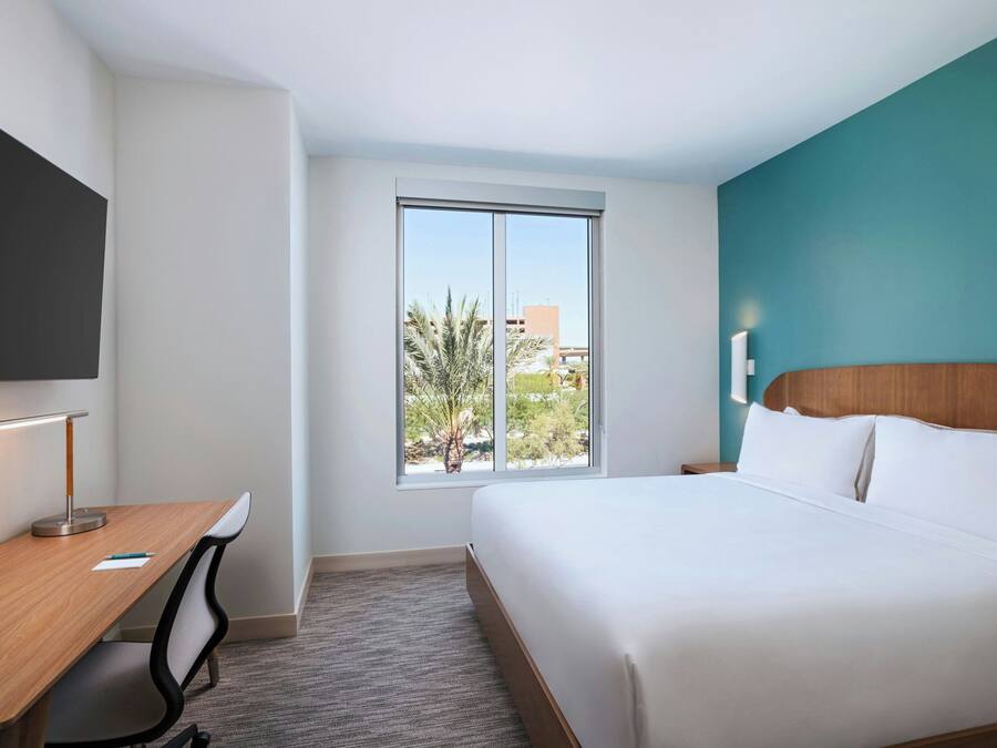 Quarto de hotel com uma cama, uma escrivaninha, uma cadeira, uma televisão fixada na parede e uma janela com vista para palmeiras e edifícios.