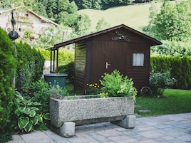 Uma cabana de madeira com telhado inclinado, uma janela e uma porta, rodeada por um jardim com um vaso de pedra e diversas plantas.