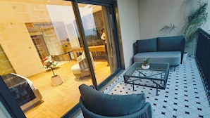 Uma sala de estar moderna com um sofá, uma mesa de centro e uma poltrona. Há uma janela grande com vista para um edifício e uma planta.