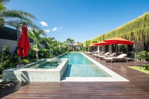 Uma piscina retangular com deck de madeira, rodeada por espreguiçadeiras e guarda-sóis vermelhos, com vegetação exuberante e um céu azul claro ao fundo.