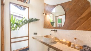 Uma casa de banho com toucador de madeira, lavatório redondo, espelho e planta em vaso.