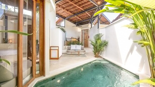 Uma área interna de piscina moderna com estrutura de vidro, teto de madeira e espaço de estar com sofá e mesa de centro.