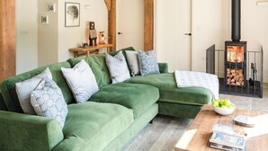 Um sofá modular verde com almofadas estampadas, uma mesa de centro de madeira com livros e uma tigela de maçãs, e uma lareira a lenha ao fundo.