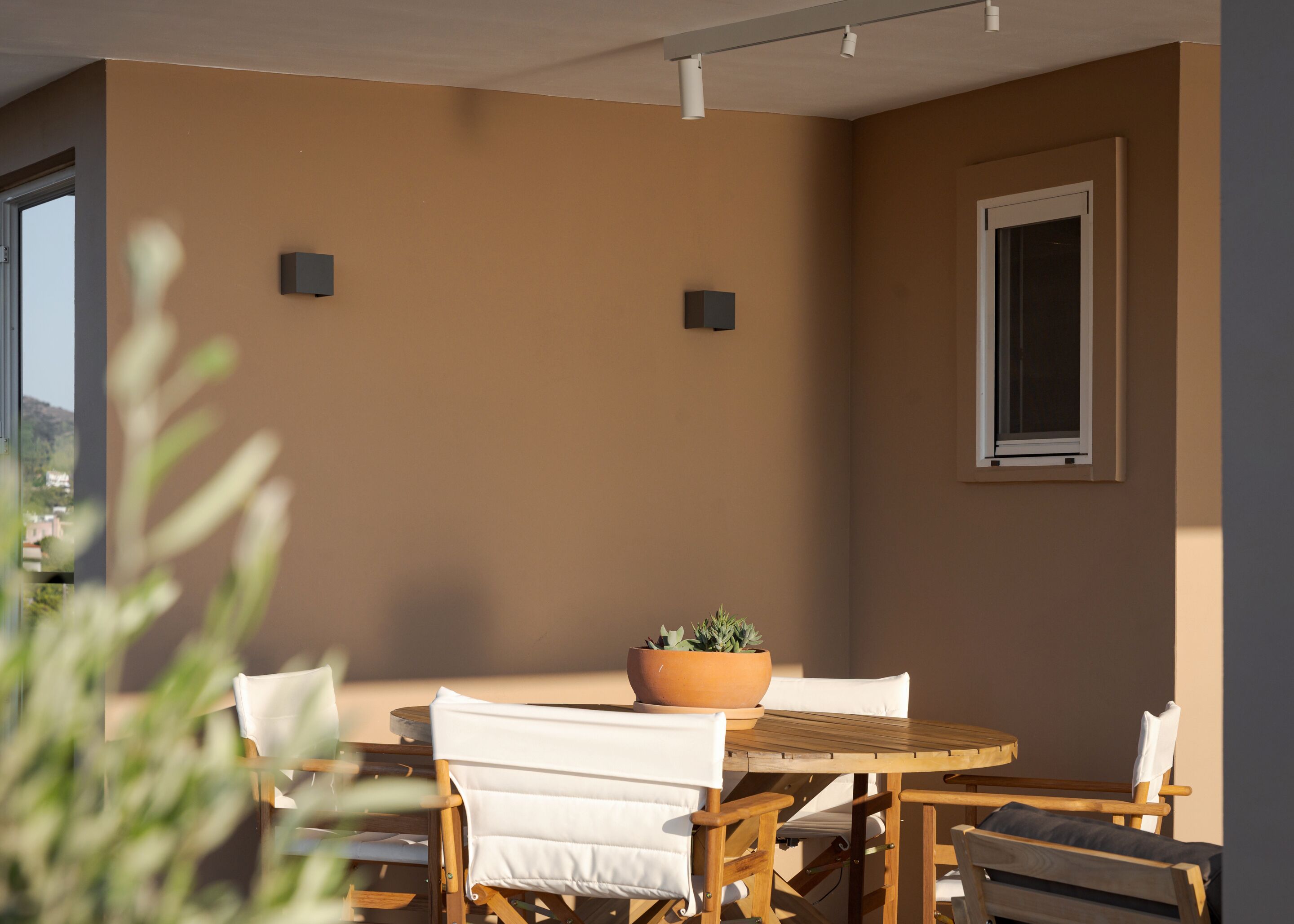 Um pátio com mesa e cadeiras de madeira, uma planta em vaso e uma janela.