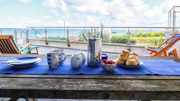Mesa posta com pratos, canecas, uma garrafa térmica, uma tigela de frutas vermelhas e doces, com vista para o oceano e cadeiras de descanso.