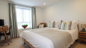 Quarto com uma cama grande, uma escrivaninha com computador, uma janela com cortinas e uma pequena planta no parapeito.
