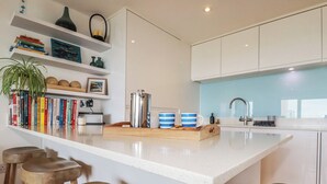 Uma cozinha moderna com ilha central, banquetas, estante com livros e objetos decorativos, e uma pia.