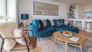 Sala de estar moderna com um sofá azul, uma poltrona macia, uma mesa de centro de madeira com frutas, e uma cozinha ao fundo.