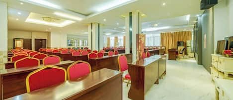 Uma sala de conferências com várias fileiras de cadeiras vermelhas, uma mesa comprida e uma televisão na parede.