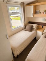 Quarto compacto com uma cama de solteiro, uma pequena mesa de cabeceira, um criado-mudo, uma janela com cortinas e uma estante com objetos decorativos.