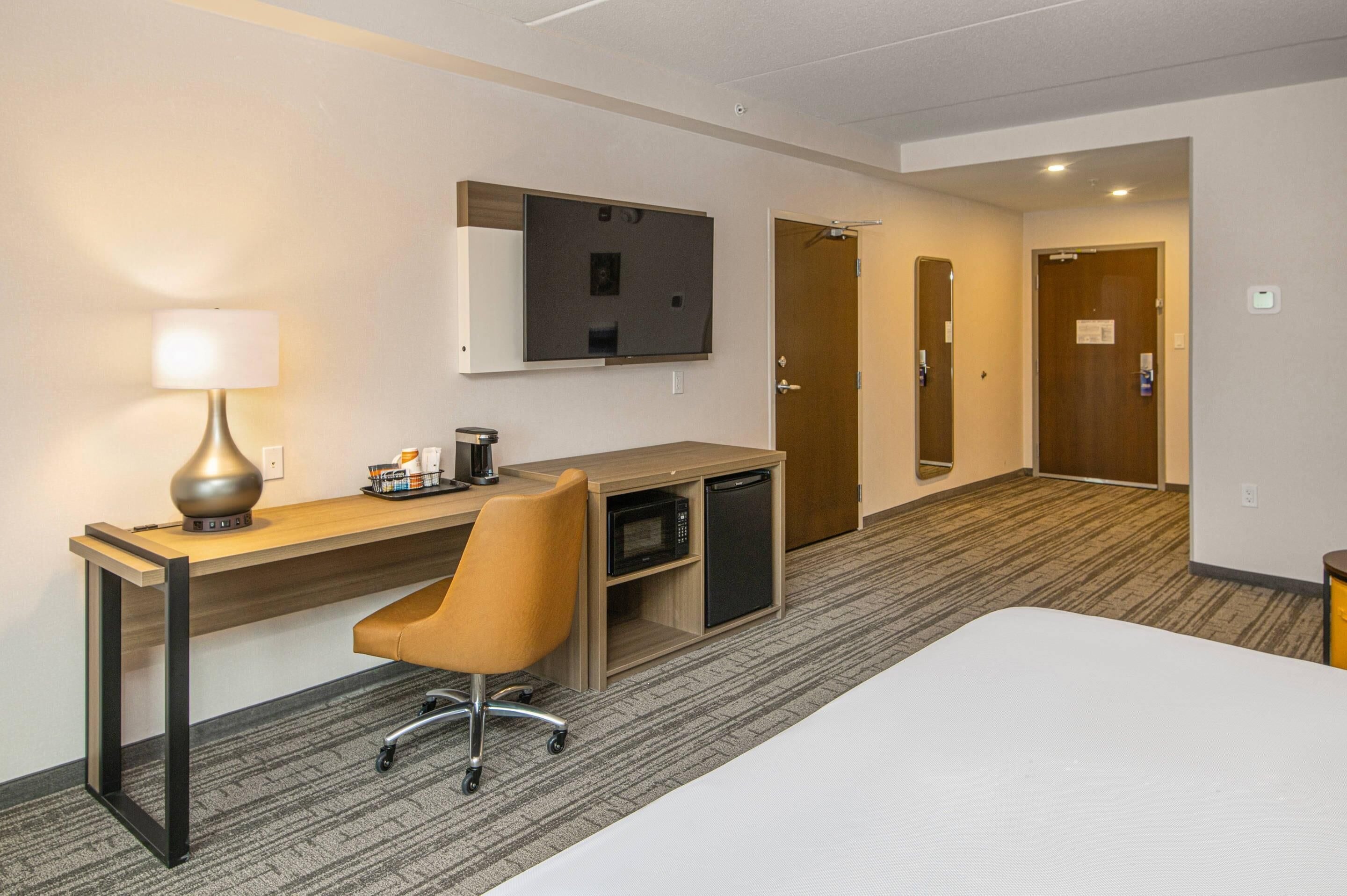Quarto de hotel com cama, escrivaninha, cadeira, micro-ondas e televisão na parede.