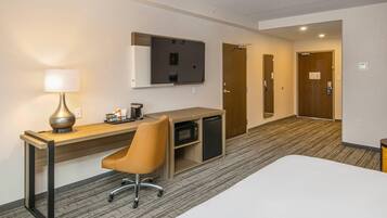 Quarto de hotel com cama, escrivaninha, cadeira, micro-ondas e televisão na parede.
