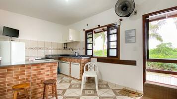 Uma cozinha compacta com piso ladrilhado, uma pequena área de refeições com banquetas, uma geladeira, uma pia e uma janela com vista para o verde.
