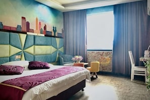 Quarto de hotel com cama, uma cadeira, uma mesinha, janela com cortinas e um mural com vista para a cidade.