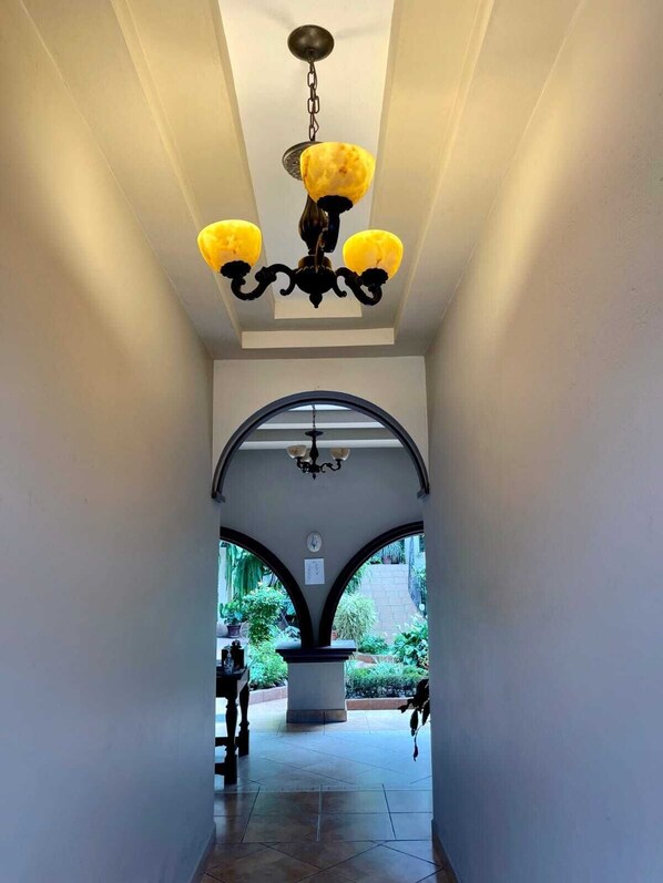 Um corredor com um candelabro e uma porta arqueada que dá para uma área externa com plantas.