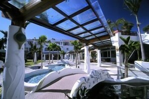 Uma área de piscina com cobertura de vidro, palmeiras e um edifício ao fundo.