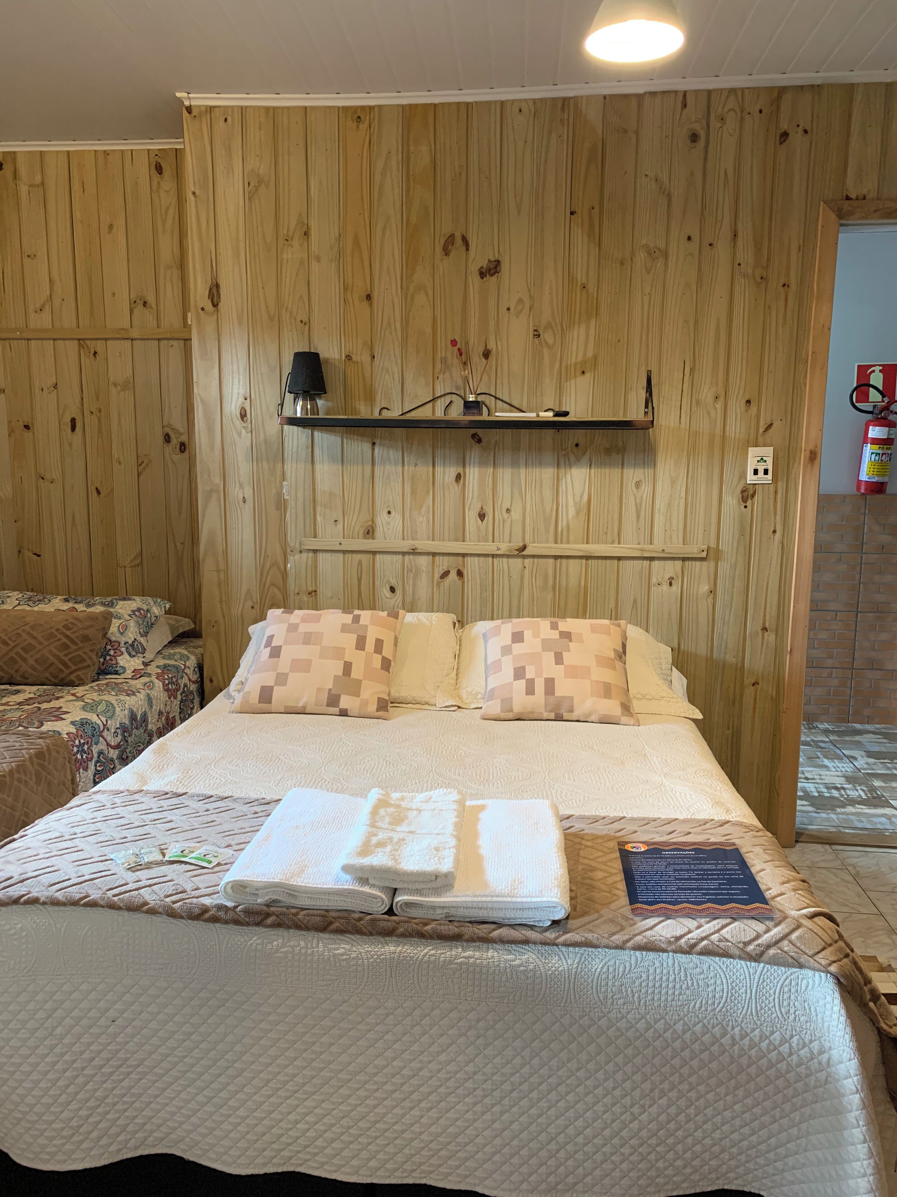Quarto de hotel com parede revestida em madeira, cama com roupa de cama estampada, mesa de cabeceira com abajur e extintor de incêndio na parede.